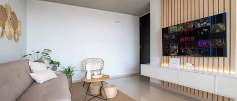 Uma sala de estar moderna com um sofá, uma mesa redonda com uma cadeira, uma televisão de tela plana grande e uma estante com objetos decorativos.