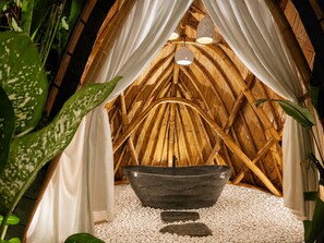 Um quarto com telhado de palha, uma bacia de pedra e pedrinhas brancas no chão.