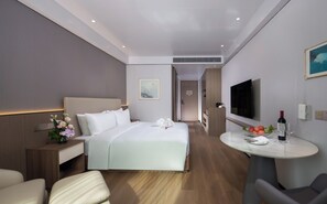 Quarto de hotel com cama, mesa de cabeceira, luminária, poltrona, televisão e uma pequena mesa de jantar com uma tigela de frutas e uma garrafa.