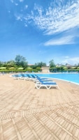 Área junto à piscina com espreguiçadeiras e guarda-sóis, céu claro e um edifício ao fundo.