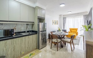 Uma cozinha moderna com micro-ondas embutido, geladeira de aço inoxidável e pia. Há uma área de jantar com mesa redonda e cadeiras, além de uma poltrona amarela.