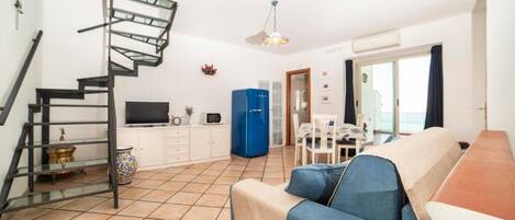 Uma sala de estar com sofá, televisão, frigorífico azul, escada e área de jantar.