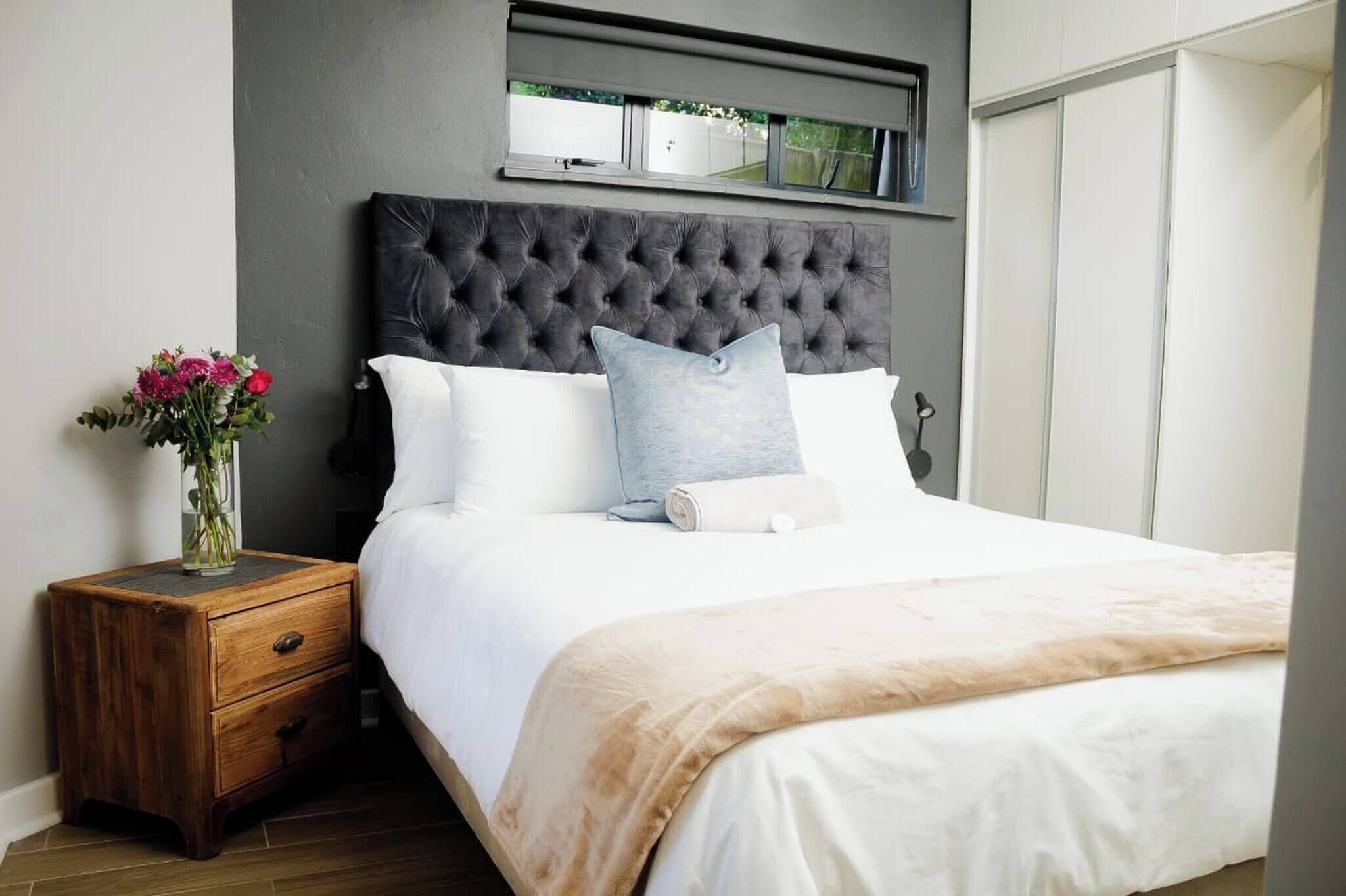 Quarto com cabeceira estofada, mesa de cabeceira de madeira com um vaso de flores e uma janela grande sobre a cama.