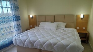 Uma cama grande com um edredom branco acolchoado, duas mesas de cabeceira com luminárias, uma cortina com padrão floral e um chão de azulejos.