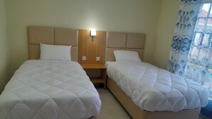 Quarto duplo com duas camas, uma mesa de cabeceira, luminária na parede e janela com cortinas estampadas.