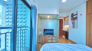 Quarto de hotel moderno com uma cama grande, televisão, escrivaninha com cadeira, pequena cozinha e varanda com vista para a cidade.