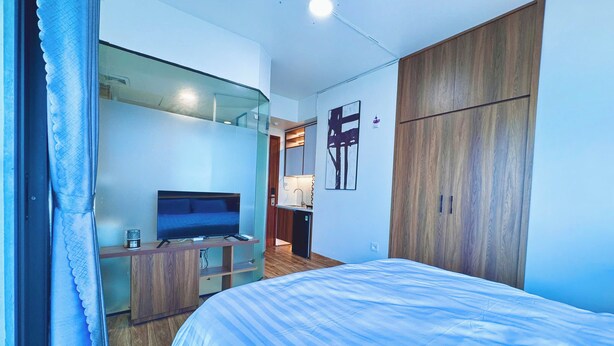 Quarto com cama, televisão, guarda-roupa de madeira e uma pequena cozinha compacta.