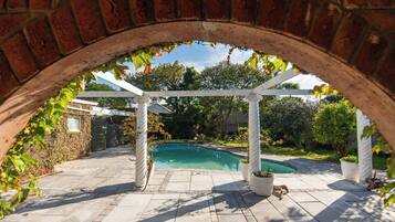 Um arco de tijolos dá acesso a uma área de piscina com um pérgula e plantas em vasos.