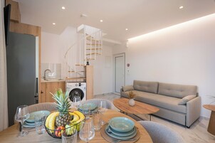 Uma sala de estar moderna com sofá, uma mesa de jantar com frutas e copos, uma cozinha com máquina de lavar e uma escada em espiral.