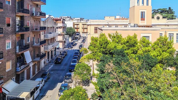 Um edifício residencial com vários andares, varandas, uma rua com carros estacionados e uma pequena área externa com mesas e cadeiras.