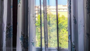 Janela com cortinas leves com padrões florais, vista para vegetação e edifícios lá fora.