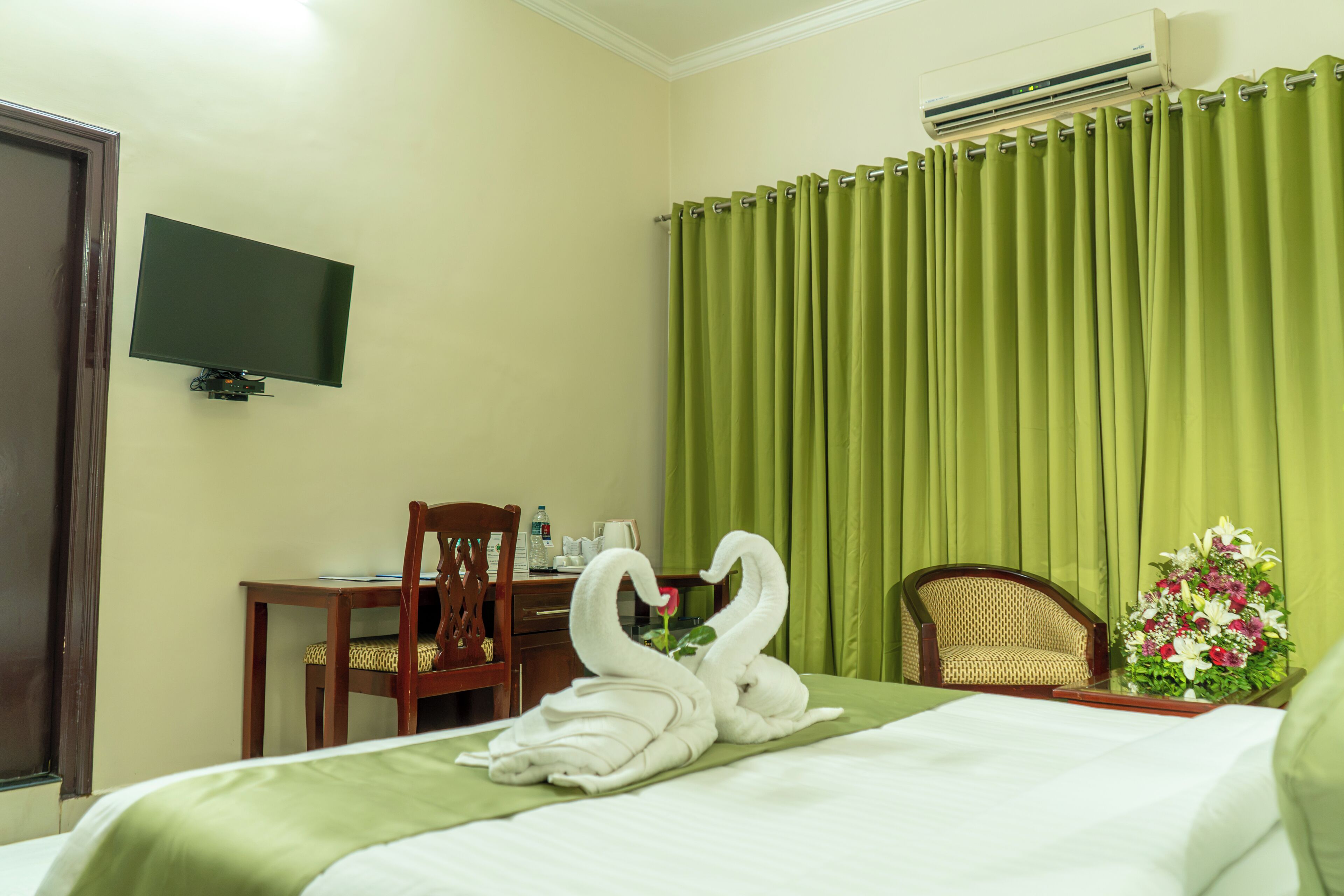 Quarto de hotel com cama, escrivaninha, cadeira, televisão e cortina verde.