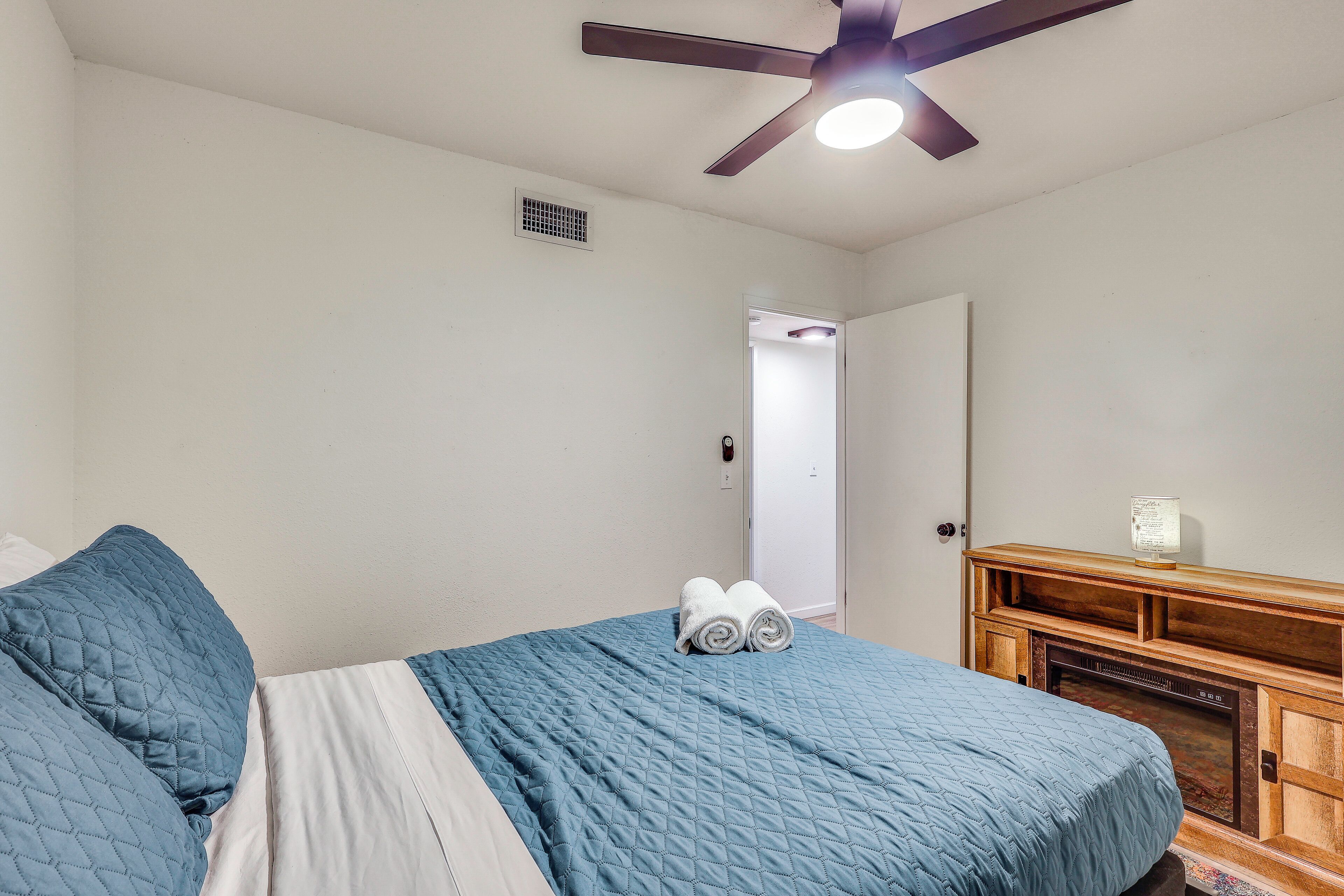 Um quarto com cama, ventilador de teto, cômoda de madeira e porta que dá para outro espaço.