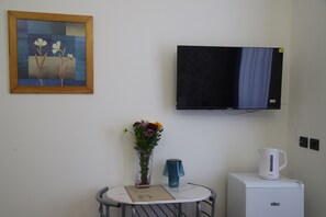 Quarto com televisão na parede, uma mesinha com um vaso de flores, chaleira e um quadro emoldurado.