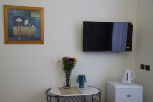 Quarto com televisão na parede, uma mesinha com um vaso de flores, chaleira e um quadro emoldurado.