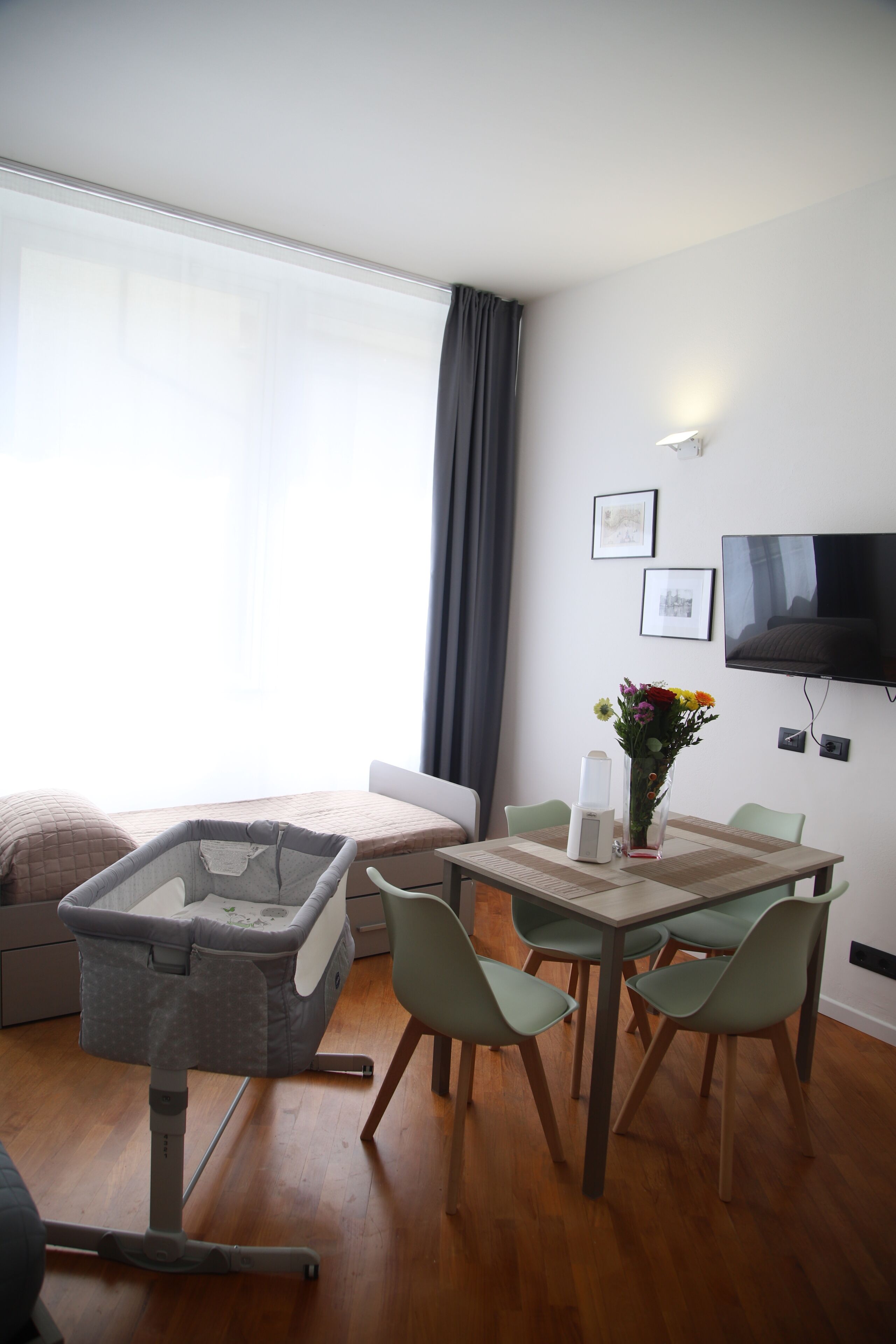 Quarto de hotel moderno com uma cama, berço para bebê, mesa de jantar com cadeiras, televisão de tela plana e uma grande janela com cortinas.