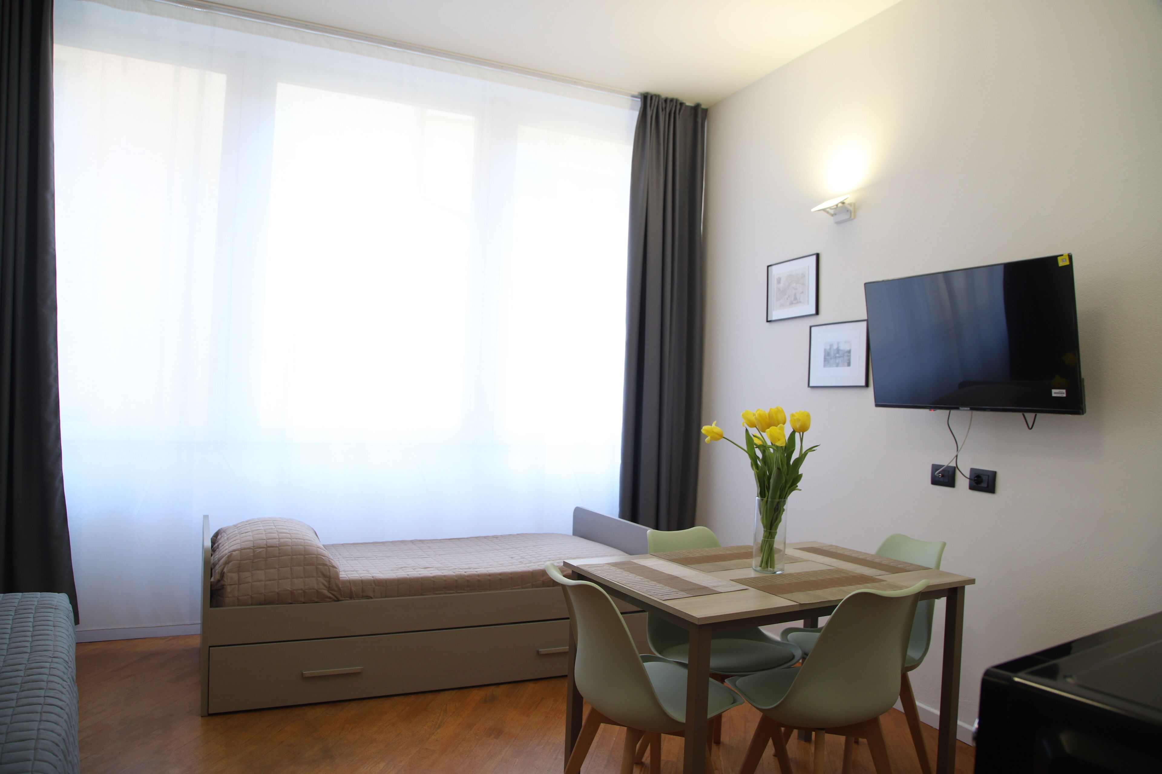 Um quarto com uma cama, uma mesa de jantar com cadeiras, uma televisão de tela plana na parede e uma janela com cortinas.