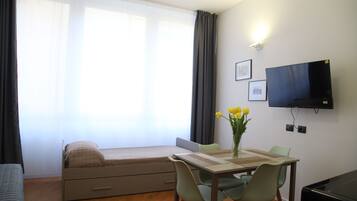 Um quarto com uma cama, uma mesa de jantar com cadeiras, uma televisão de tela plana na parede e uma janela com cortinas.
