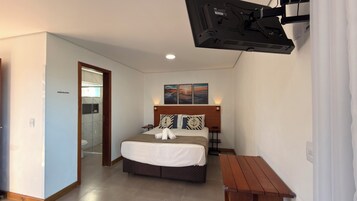 Quarto de hotel com uma cama, mesas de cabeceira, um banco de madeira, área de chuveiro e televisão na parede.