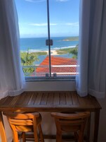 Uma mesa de madeira com duas cadeiras, uma janela com cortinas brancas e vista para o oceano e um prédio com telhado vermelho.