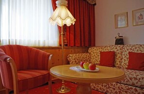 Um quarto com uma poltrona listrada, um sofá estampado com flores, uma mesa redonda com um prato de frutas, um abajur e uma janela com cortinas.