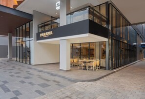 Um edifício moderno com fachada de vidro, um letreiro com a inscrição 'PARAMAX' e área externa com mesas e cadeiras.
