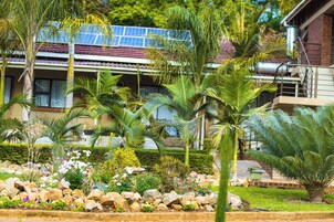 Uma casa com painéis solares, fachada de tijolos e um jardim bem cuidado, com diversas plantas e pedras.
