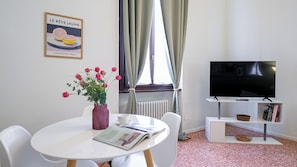Um quarto com uma mesa de jantar, um vaso de flores, um quadro na parede, um radiador, uma janela com cortinas, uma televisão sobre um suporte e uma estante de livros.