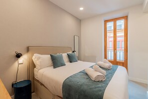 Cama bem arrumada com almofadas, abajur e janela com vista para edifícios.