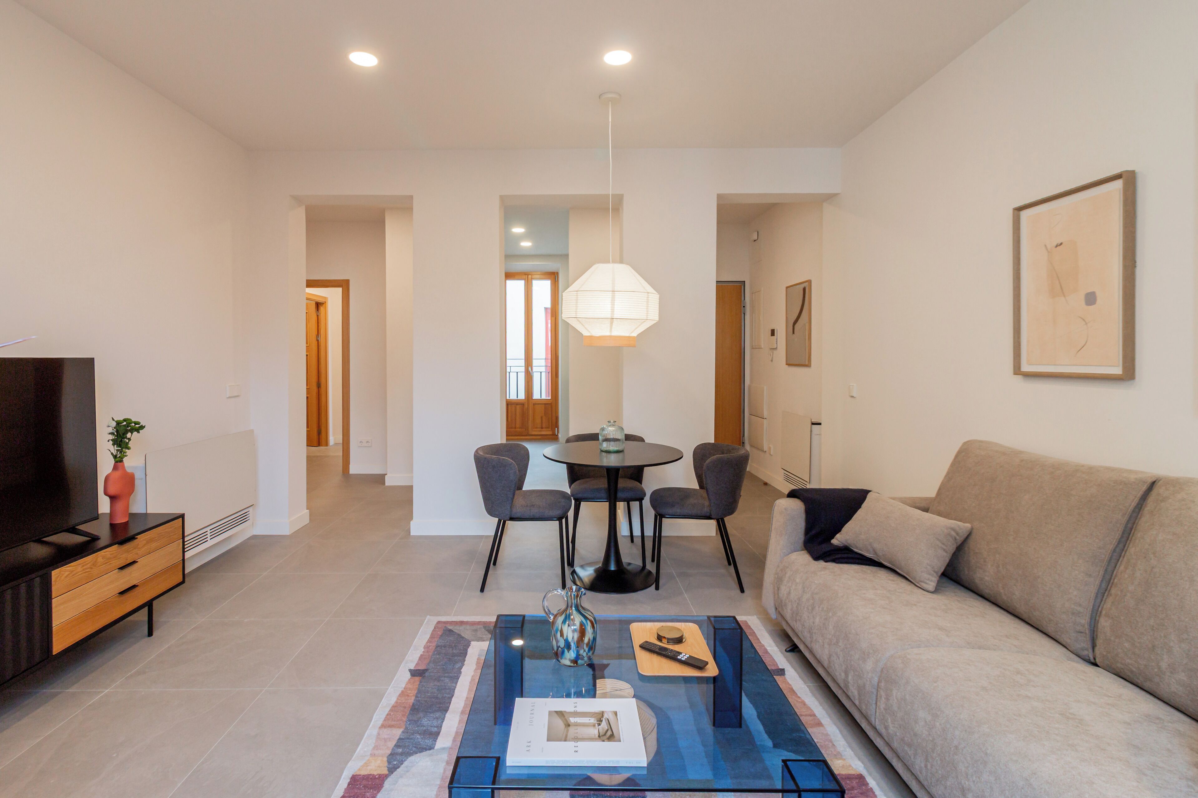 A modern living room with a sofa, a coffee table with a book and remote, a TV on a stand, and a dining area with chairs and a table.