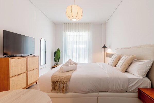 Quarto com cama, televisão, mesa de cabeceira, abajur e janela com cortinas.