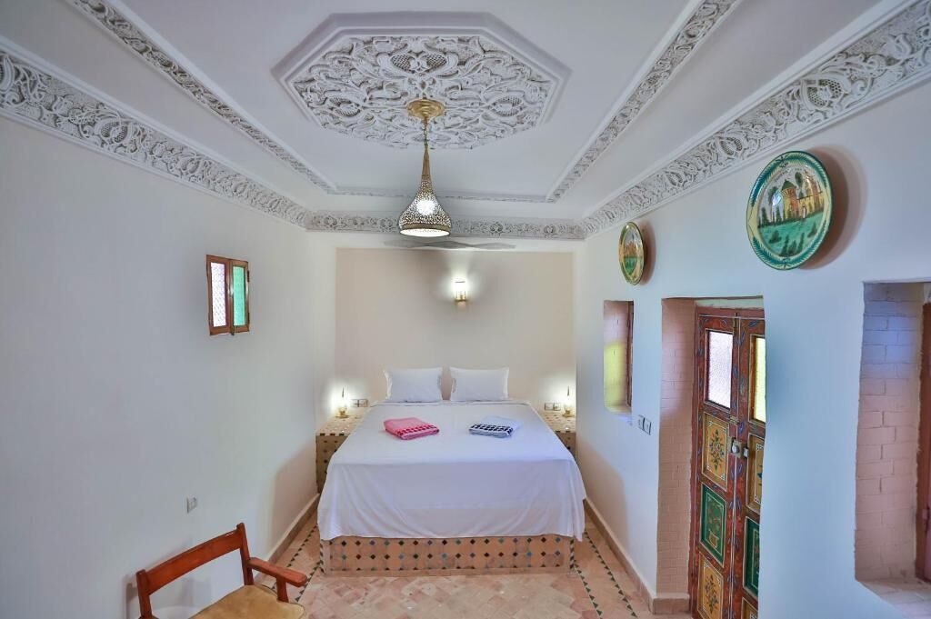 Quarto com cama, teto decorativo, luminárias fixadas na parede e porta com detalhes elaborados.
