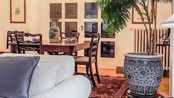 Uma sala de estar com um sofá branco, mesa de jantar, cadeiras, um candelabro, uma grande planta em vaso e um vaso decorativo.