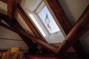 Quarto com teto inclinado, vigas de madeira, claraboia e vista para um campanário de igreja.