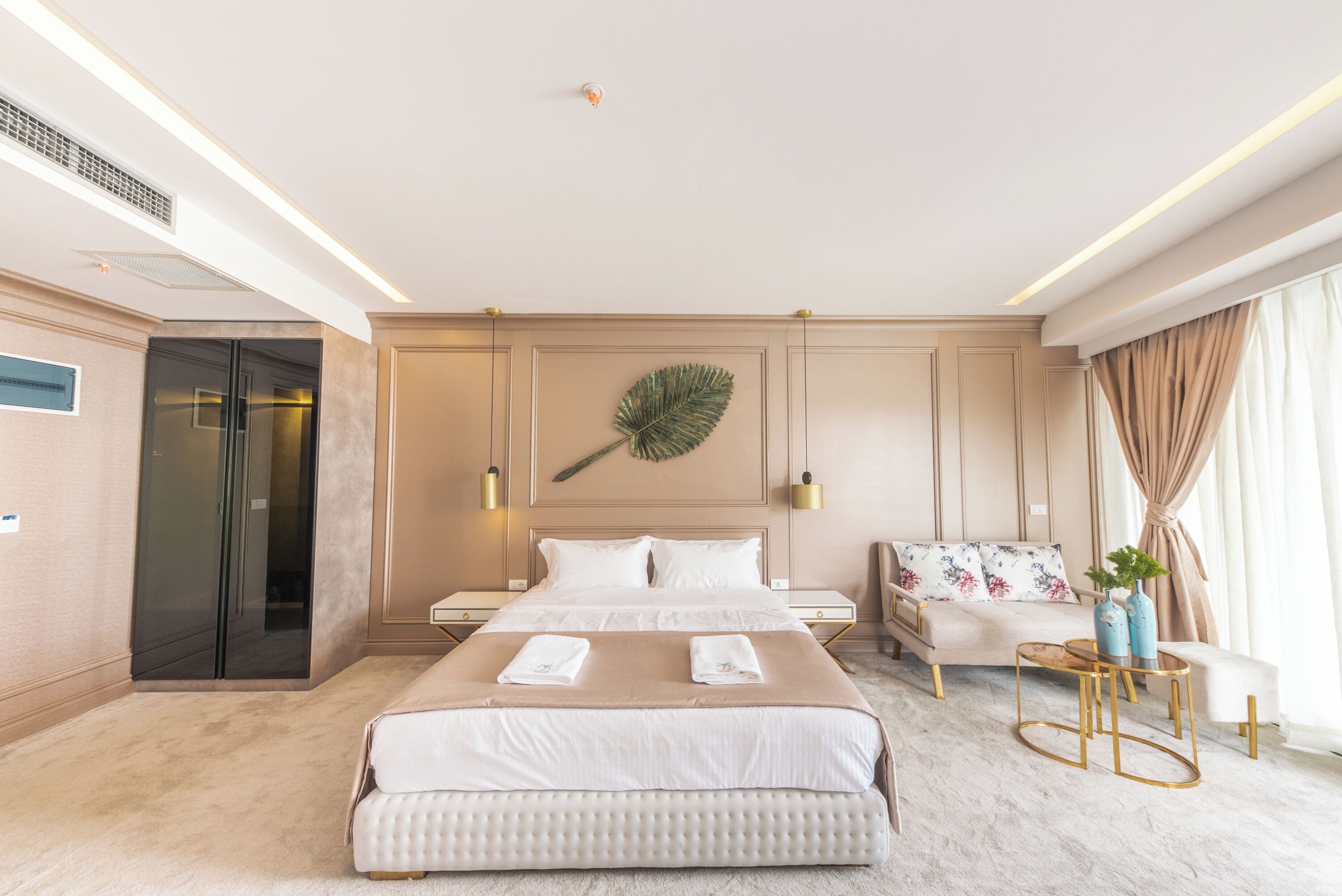 Quarto de hotel com uma cama grande, um sofá, uma mesinha e uma folha decorativa na parede.