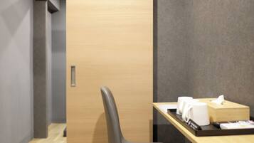 Quarto de hotel com porta de madeira, uma escrivaninha com caixa de lenços, um controle remoto e uma cadeira.