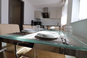 Uma área de refeições com mesa de vidro, cadeiras e cozinha ao fundo.