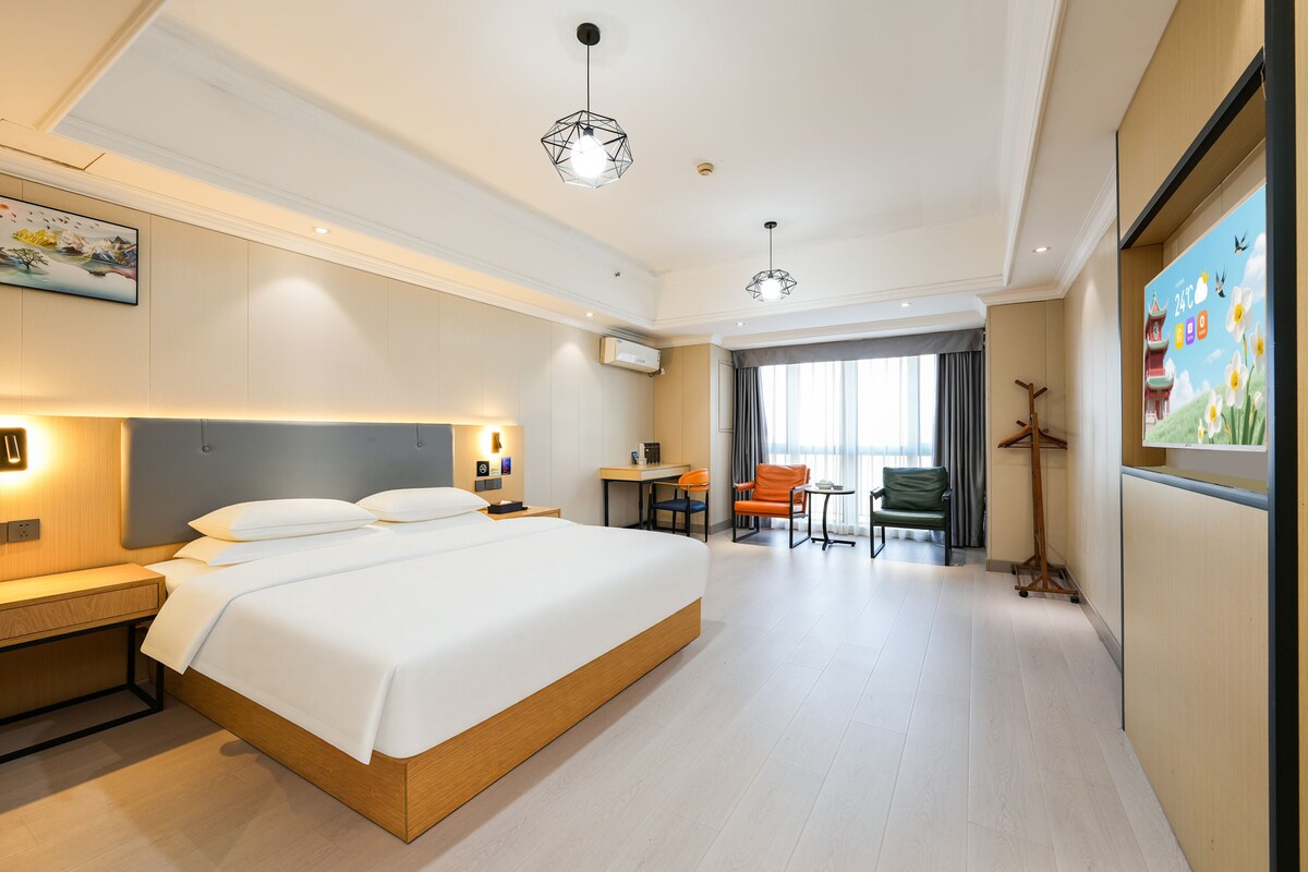Quarto de hotel com uma cama grande, mesas de cabeceira, uma escrivaninha e uma cadeira. Há uma janela com cortinas, um quadro na parede e um lustre.