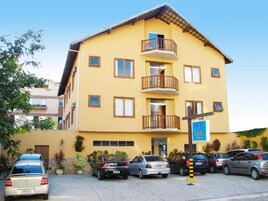Um edifício amarelo de vários andares com uma varanda no segundo andar, um estacionamento com vários carros e uma placa indicando um hotel.