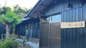 Um edifício tradicional japonês com fachada de madeira, uma placa com caracteres japoneses e uma árvore em primeiro plano.