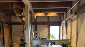 Uma área de refeições rústica com uma mesa de madeira e cadeiras pretas, uma cozinha ao fundo e uma janela grande.
