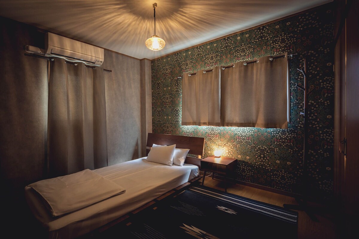 Quarto com cama, mesa de cabeceira, luminária na parede e janela com cortinas.