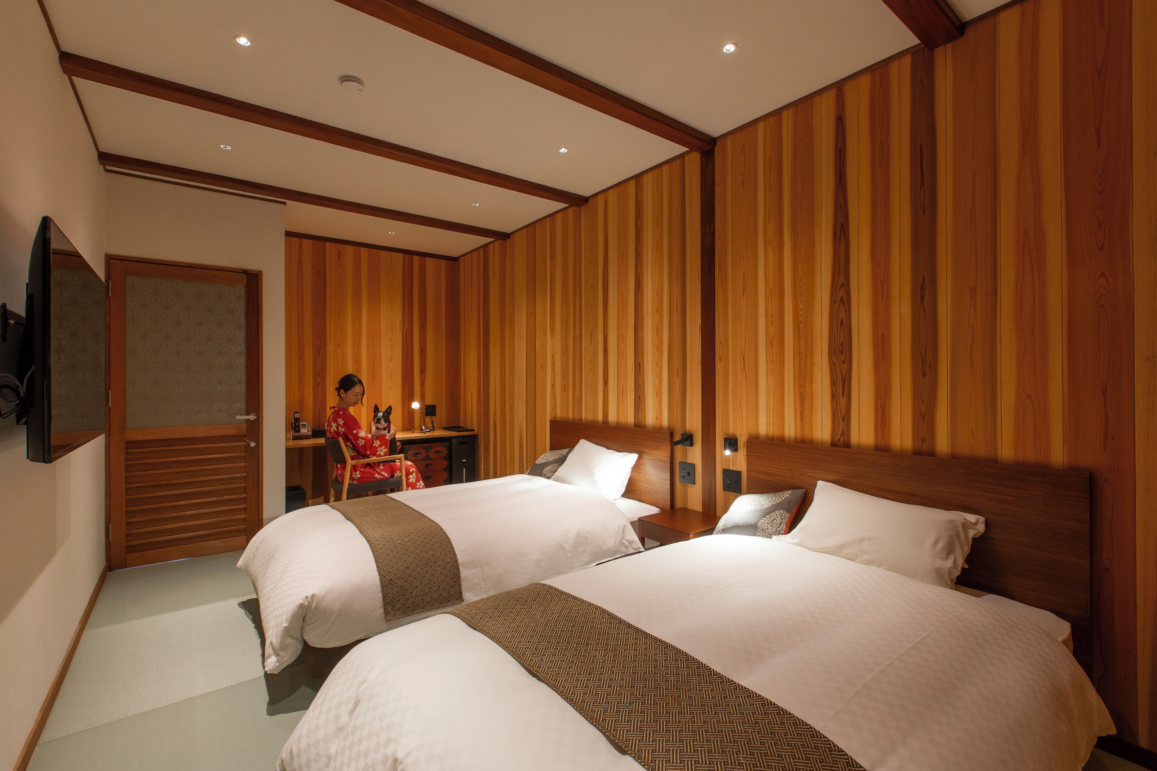 Quarto de hotel com duas camas, painéis de madeira, uma escrivaninha com cadeira e uma televisão fixada na parede.