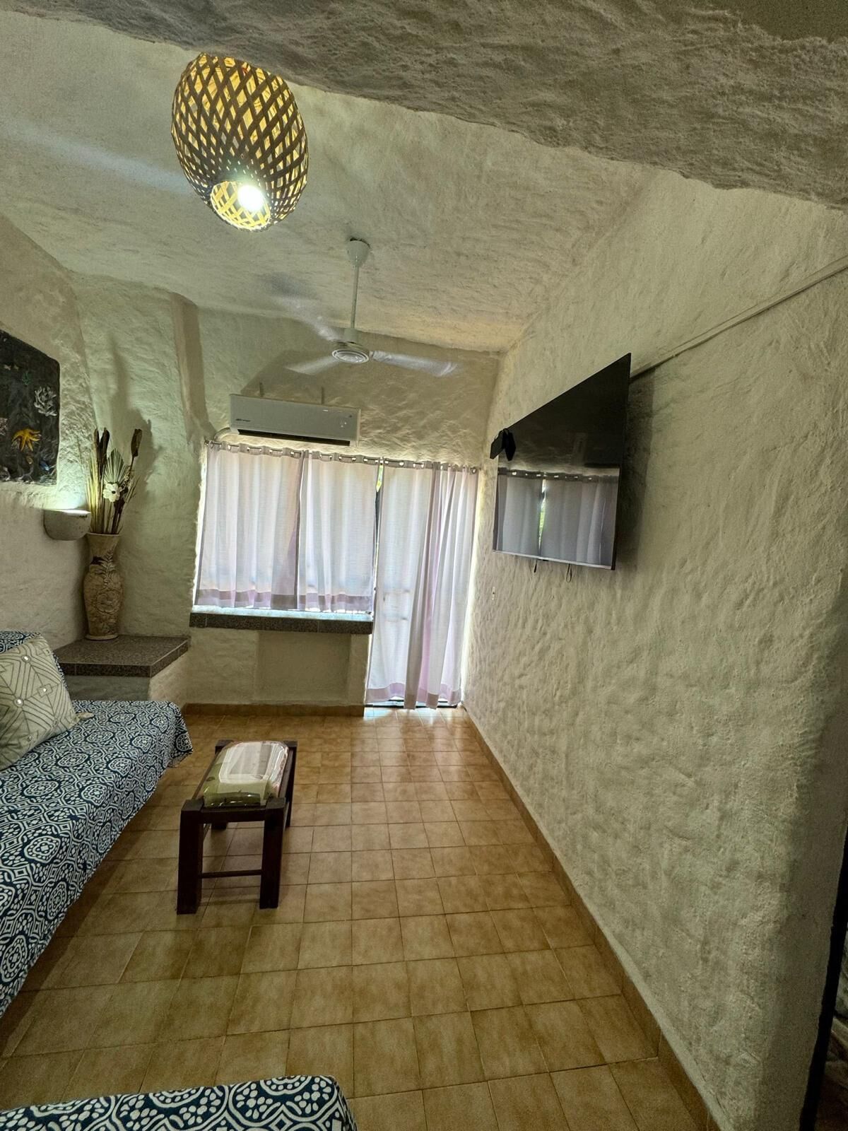Uma sala de estar com um sofá estampado, uma pequena mesa de madeira, um ventilador de teto, uma televisão fixada na parede e uma janela com cortinas leves.