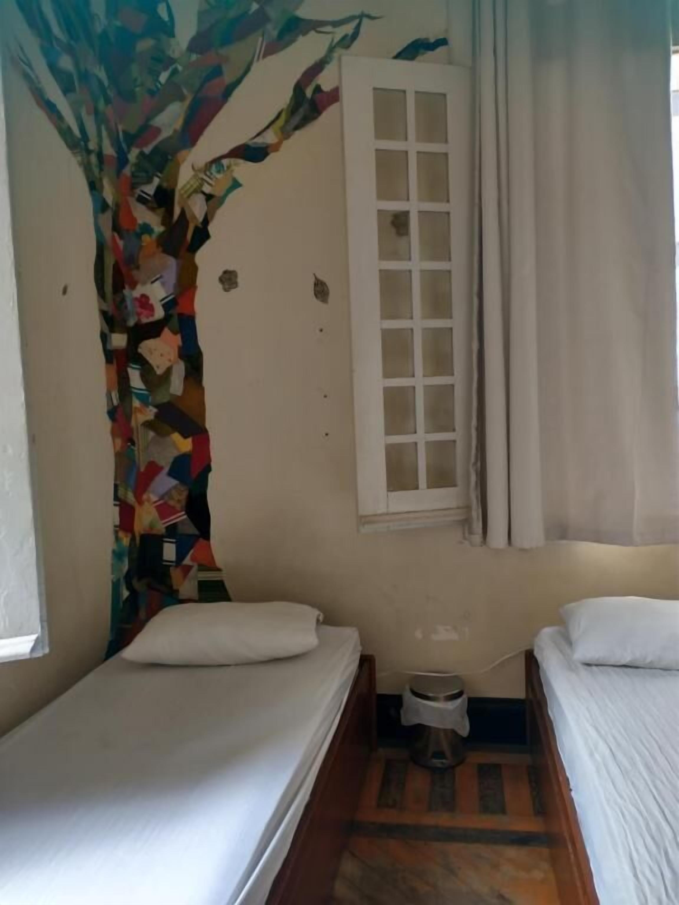Um pequeno alojamento de um único quarto, com cama, janela com cortinas e um mural na parede.
