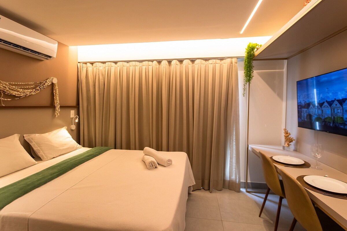 Quarto de hotel com cama, cortinas, televisão, mesa de jantar com cadeiras e ar condicionado.