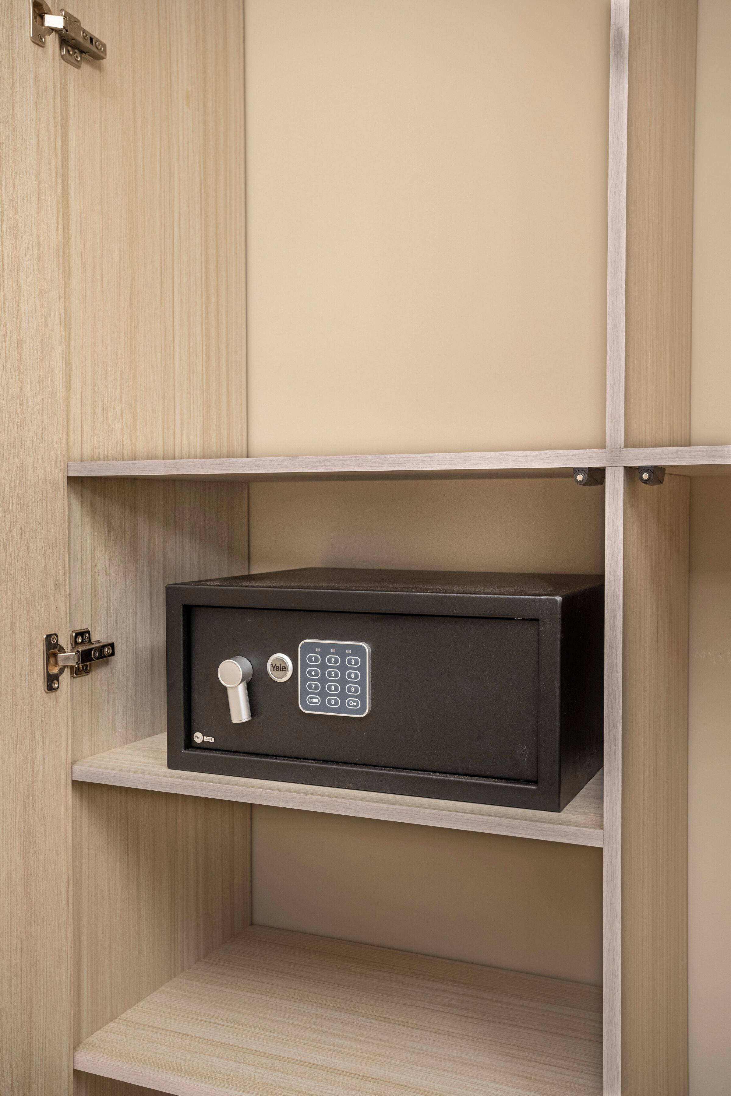 Um cofre preto com teclado numérico e fechadura, fixado numa prateleira de madeira num quarto com paredes e portas de madeira.