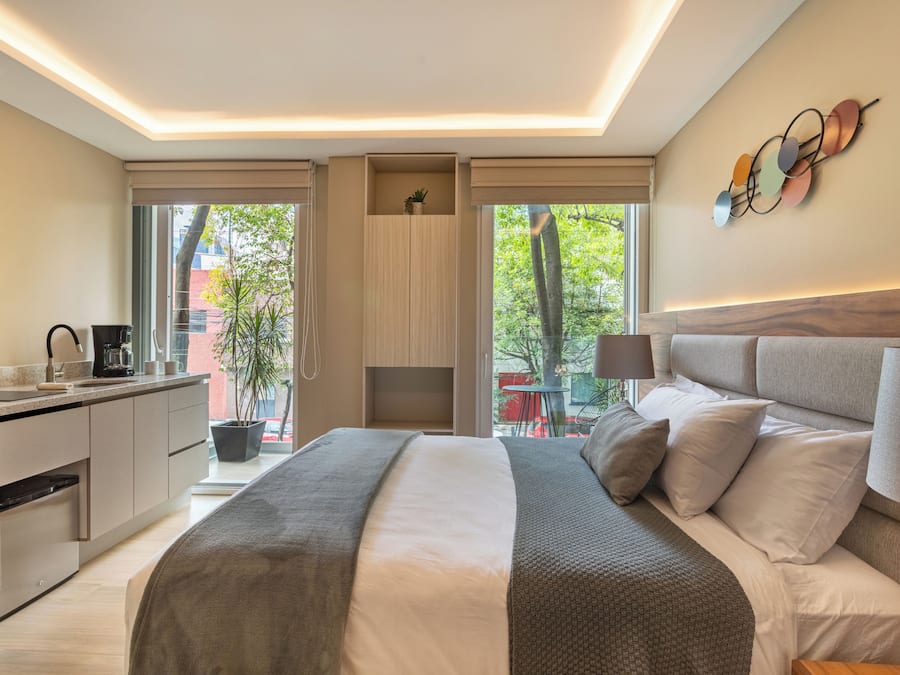 Quarto de hotel moderno com uma cama, uma escrivaninha com máquina de café, uma planta em vaso e vista exterior.