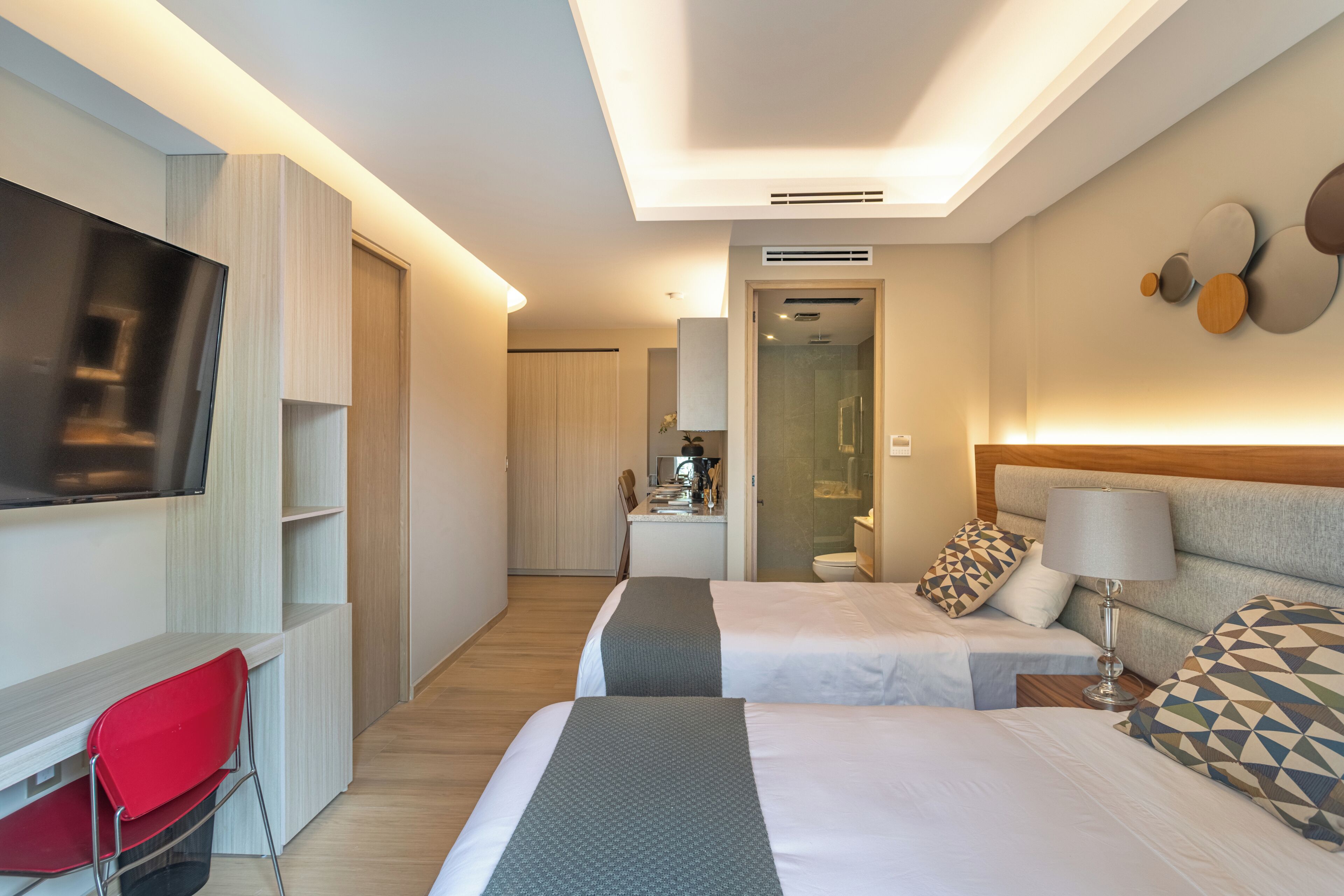 Quarto de hotel com duas camas, uma escrivaninha com uma cadeira vermelha, uma televisão e um banheiro visível ao fundo.