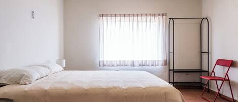 Um quarto com uma cama, uma janela com cortinas leves, uma cadeira vermelha e uma estante de metal.